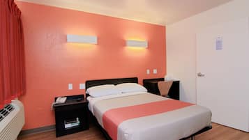 Quarto de hotel com cama, mesa de cabeceira, escrivaninha, cadeira, porta e luminária fixada na parede.