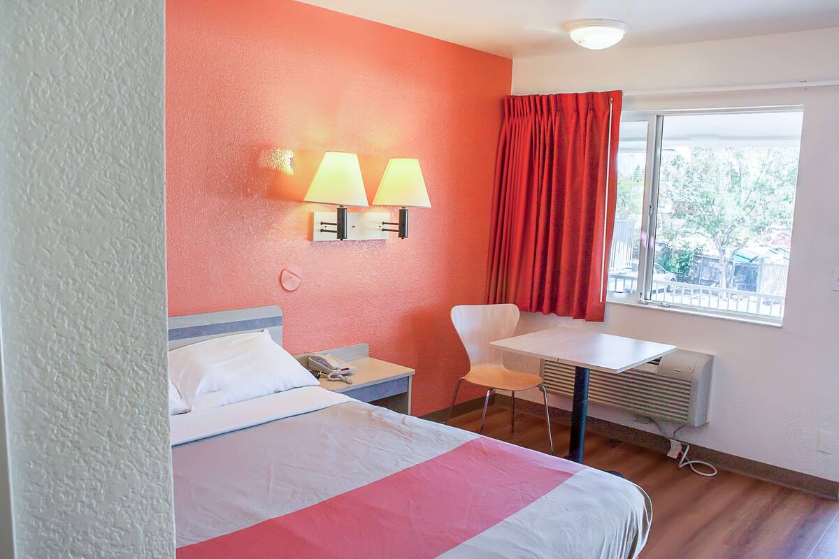 Quarto de hotel com uma cama, uma escrivaninha, duas luminárias fixadas na parede, uma cadeira, uma janela com cortinas vermelhas e vista para árvores lá fora.