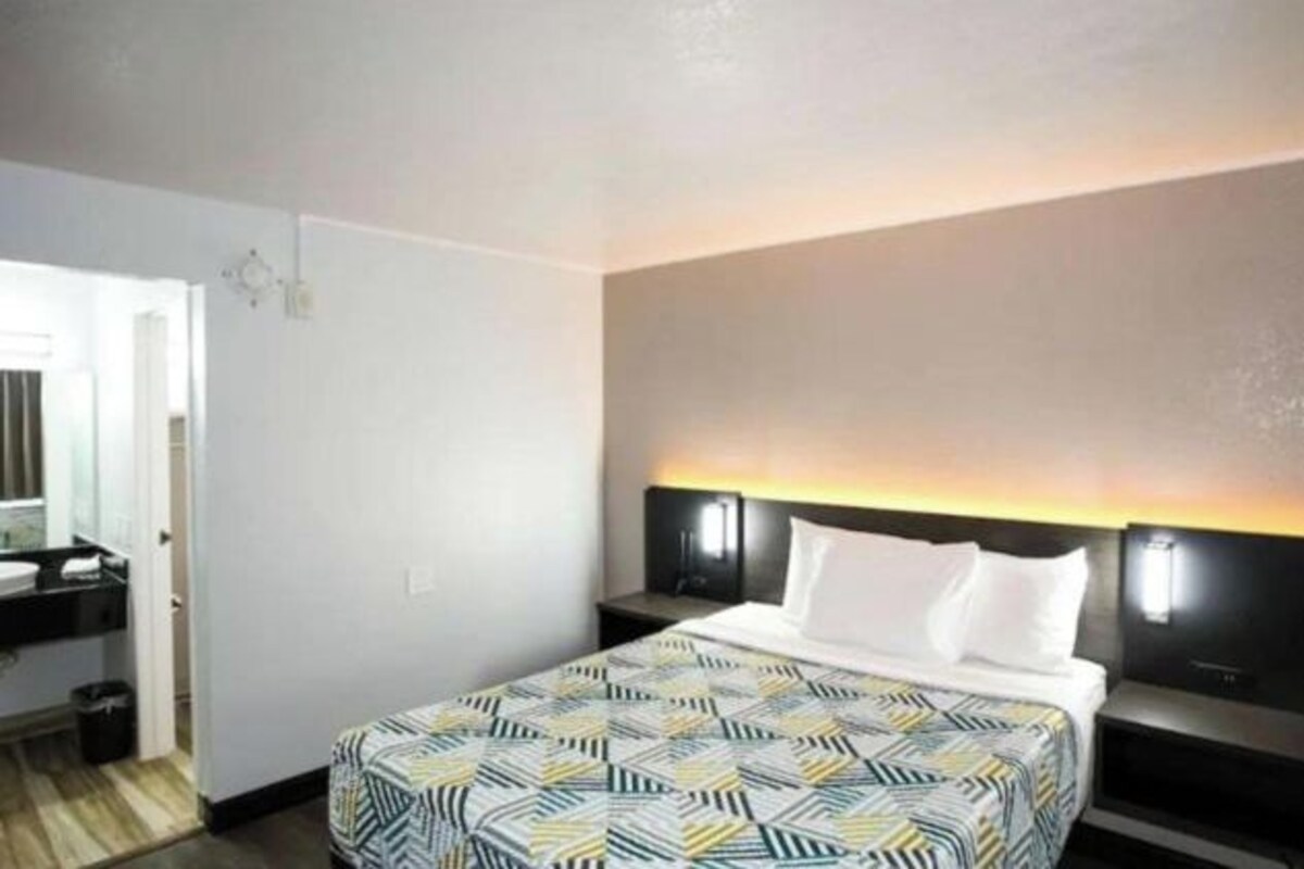 Quarto de hotel com cama, mesa de cabeceira, luminária na parede e banheiro com pia e espelho.