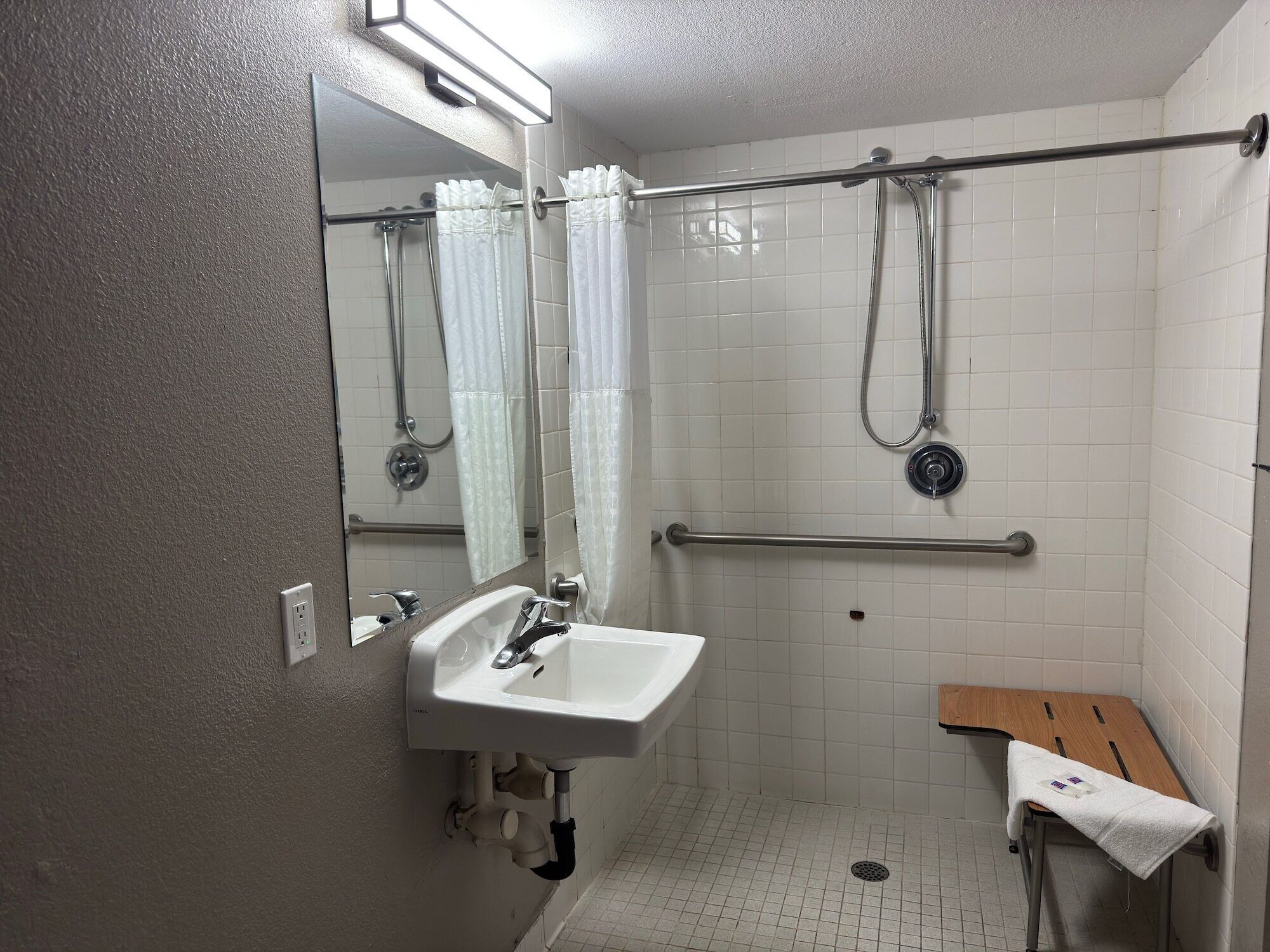 Casa de banho com lavatório branco, espelho e zona de duche com barra de apoio.
