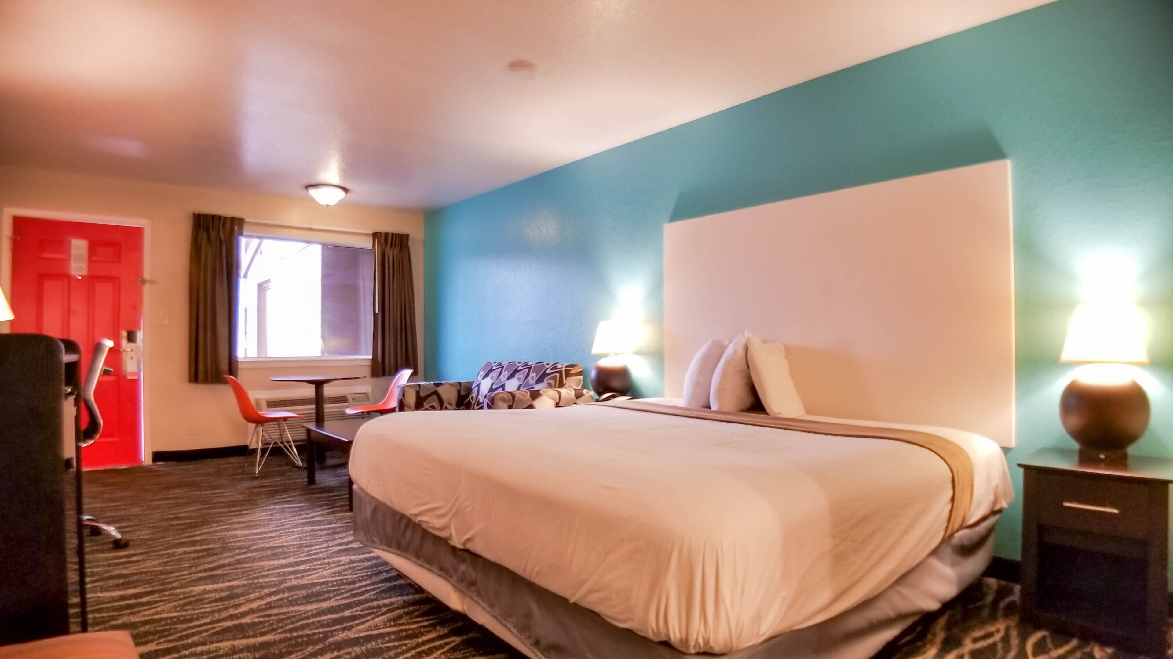 Quarto de hotel com cama grande, uma escrivaninha, duas cadeiras, uma porta vermelha e uma janela com cortinas.
