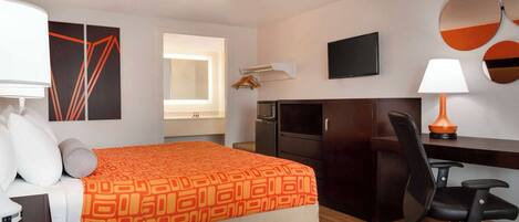 Quarto de hotel com cama, escrivaninha, cadeira e televisão na parede.