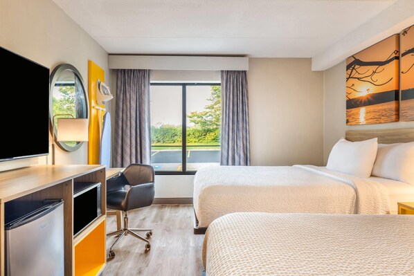Quarto de hotel com cama, escrivaninha com televisão, uma cadeira e janela com cortinas.
