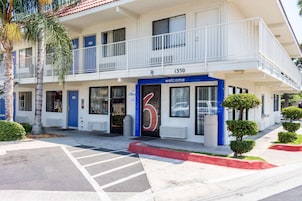 Um motel de dois andares com fachada azul e branca, numerado 1350, com um grande letreiro de entrada com o número '6', uma varanda no andar superior e um estacionamento à frente.
