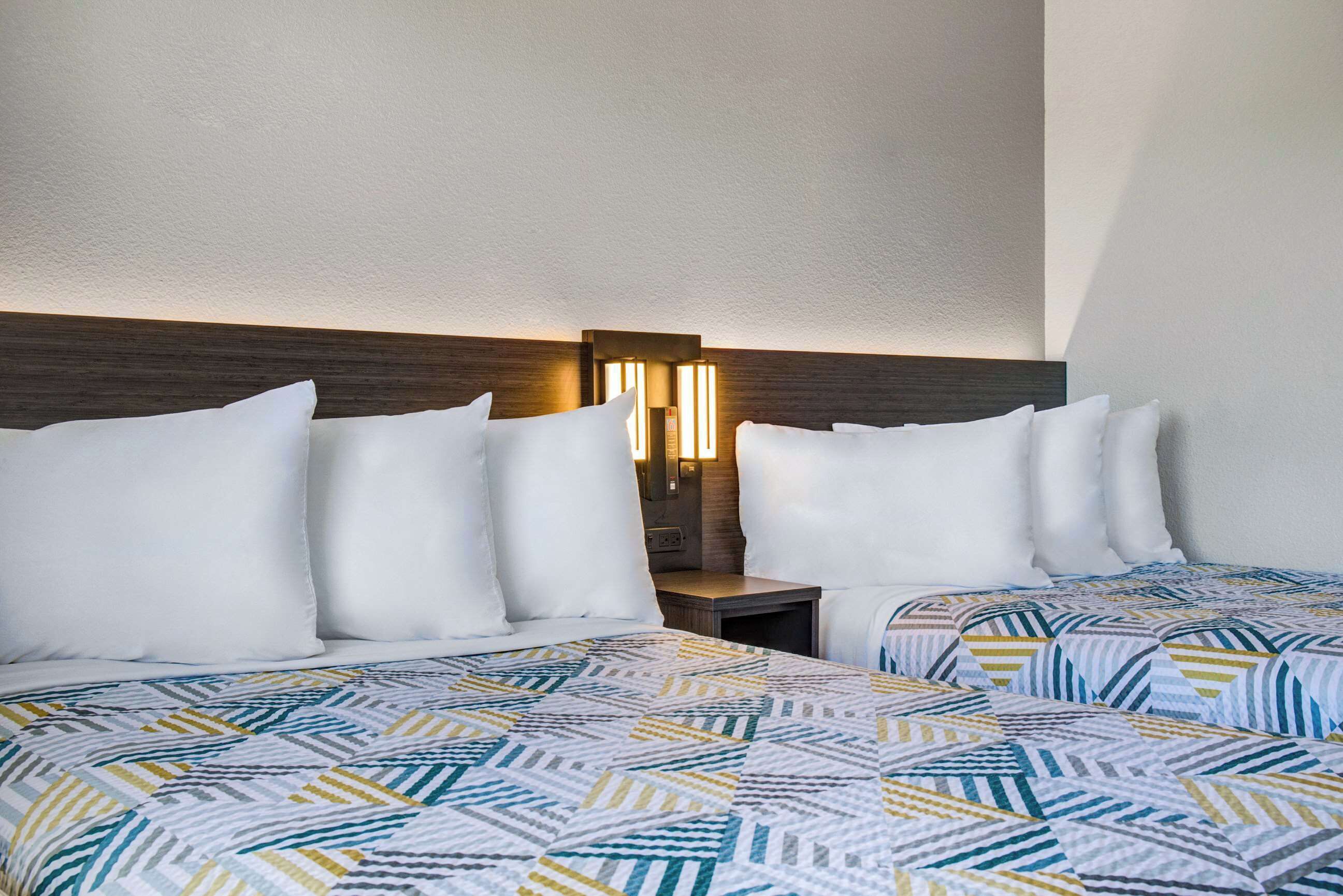 Quarto de hotel com duas camas, cada uma com roupa de cama branca e um edredom estampado. Há uma mesa de cabeceira com abajur entre as camas, e uma cabeceira de madeira.