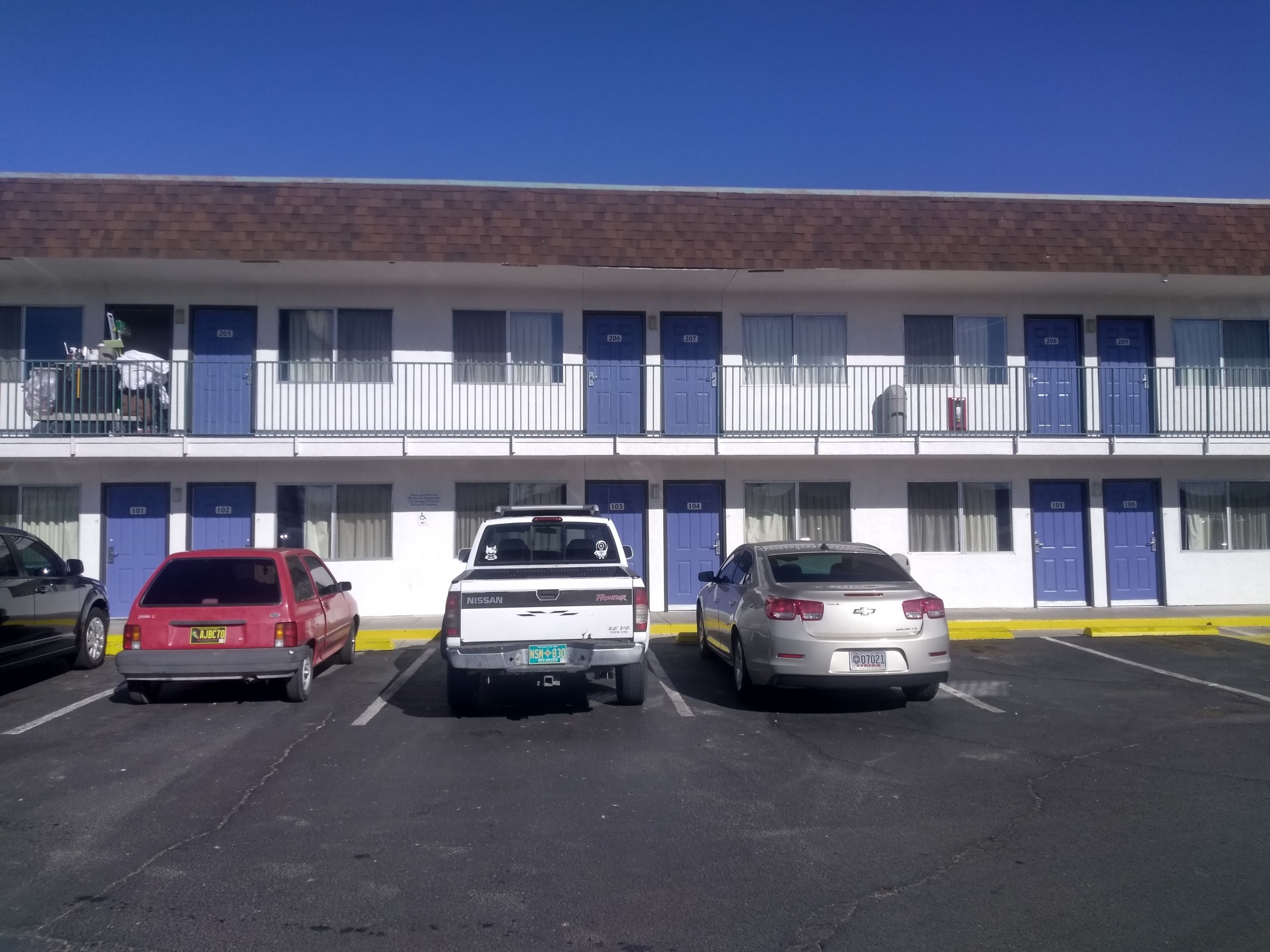 Um motel com uma fileira de quartos, cada um com uma porta azul e uma varanda. Três carros estão estacionados no estacionamento.