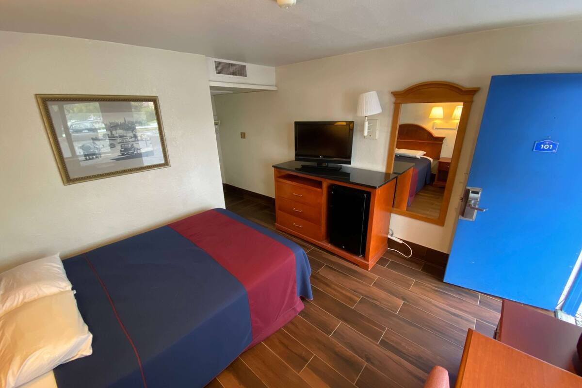 Quarto de hotel com cama, escrivaninha com televisão, espelho e porta com o número 101.