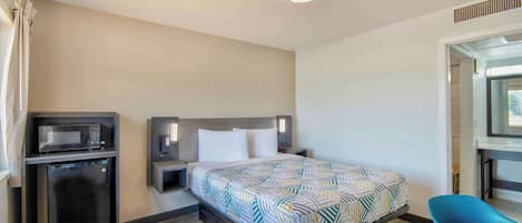 Quarto de hotel com cama, micro-ondas, mini-geladeira, mesa de cabeceira e uma cadeira azul.