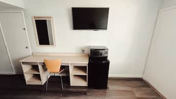 Quarto pequeno e compacto com uma escrivaninha de madeira, uma cadeira, televisão fixada na parede e um frigorífico preto.