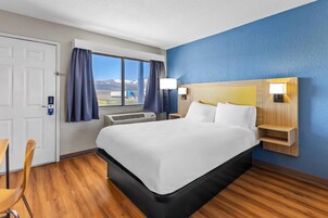 Quarto de hotel com cama, escrivaninha, cadeira, janela com cortinas e vista para montanhas.