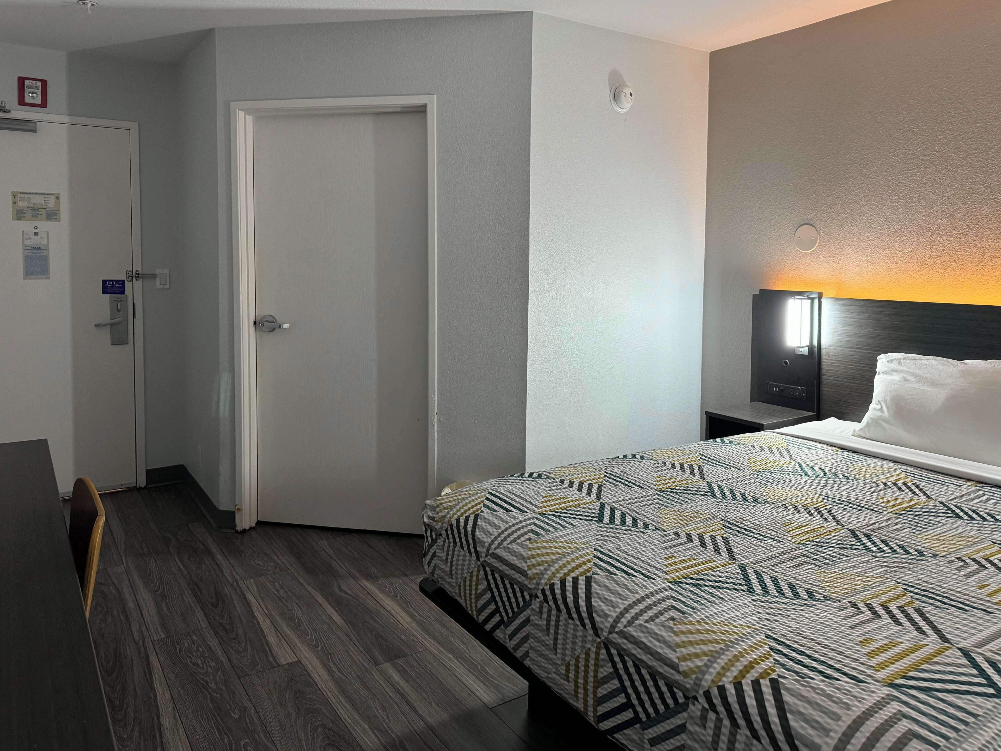 Quarto de hotel com cama, mesa de cabeceira com abajur, porta e pequena escrivaninha com cadeira.