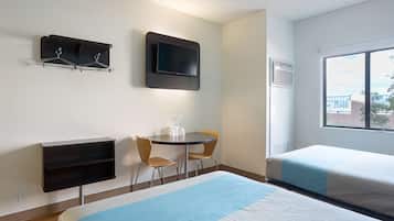 Quarto de hotel com duas camas, televisão fixada na parede, uma mesinha com cadeiras e janela com vista.