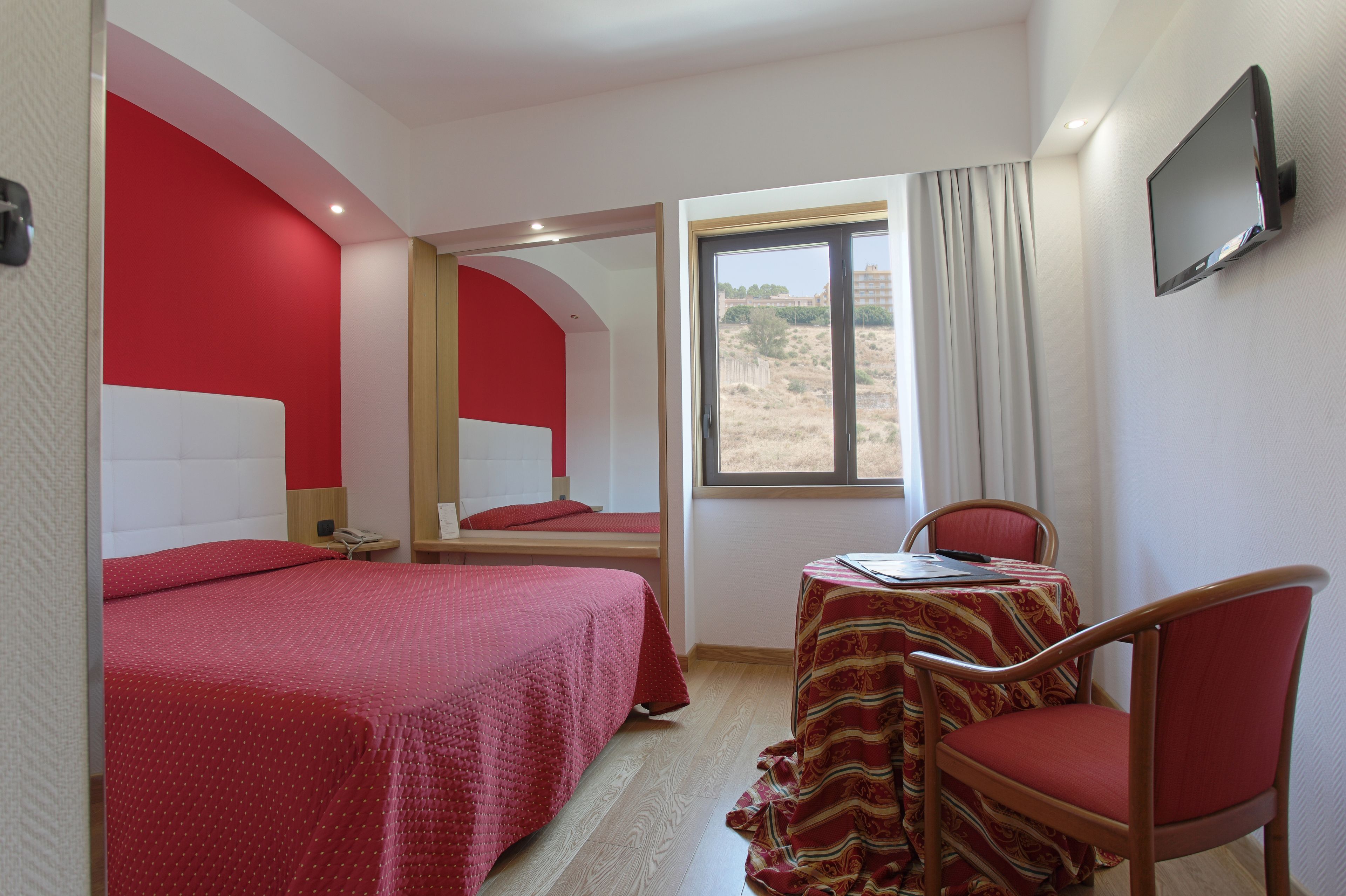 Quarto de hotel com uma cama, uma mesa com cadeira, uma televisão na parede e uma janela com cortinas.