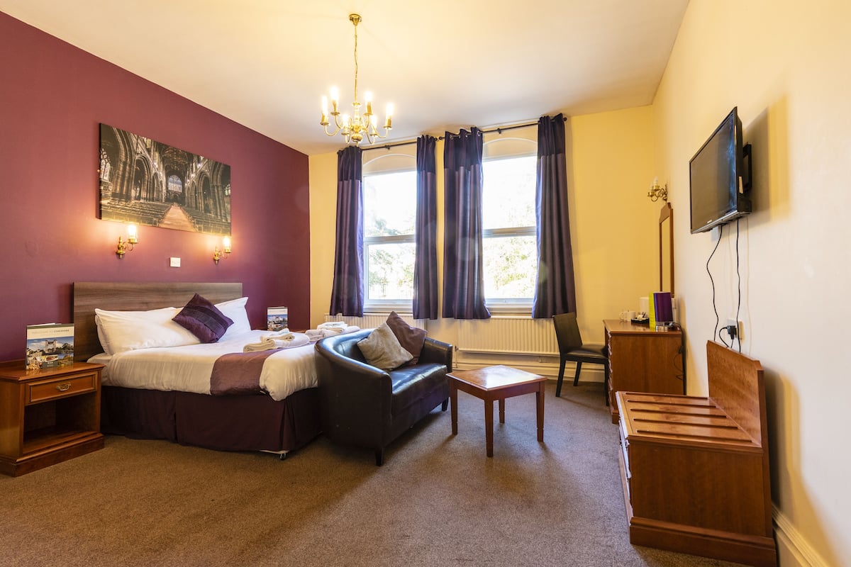 Quarto de hotel com uma cama grande, uma mesa, uma cadeira, uma TV e uma janela com cortinas.