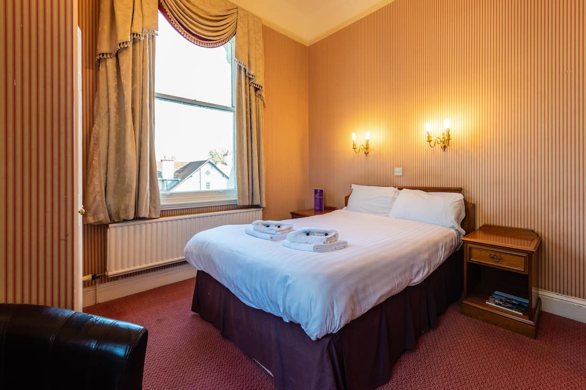 Quarto de hotel com cama, mesa de cabeceira, cadeira e janela com cortinas.
