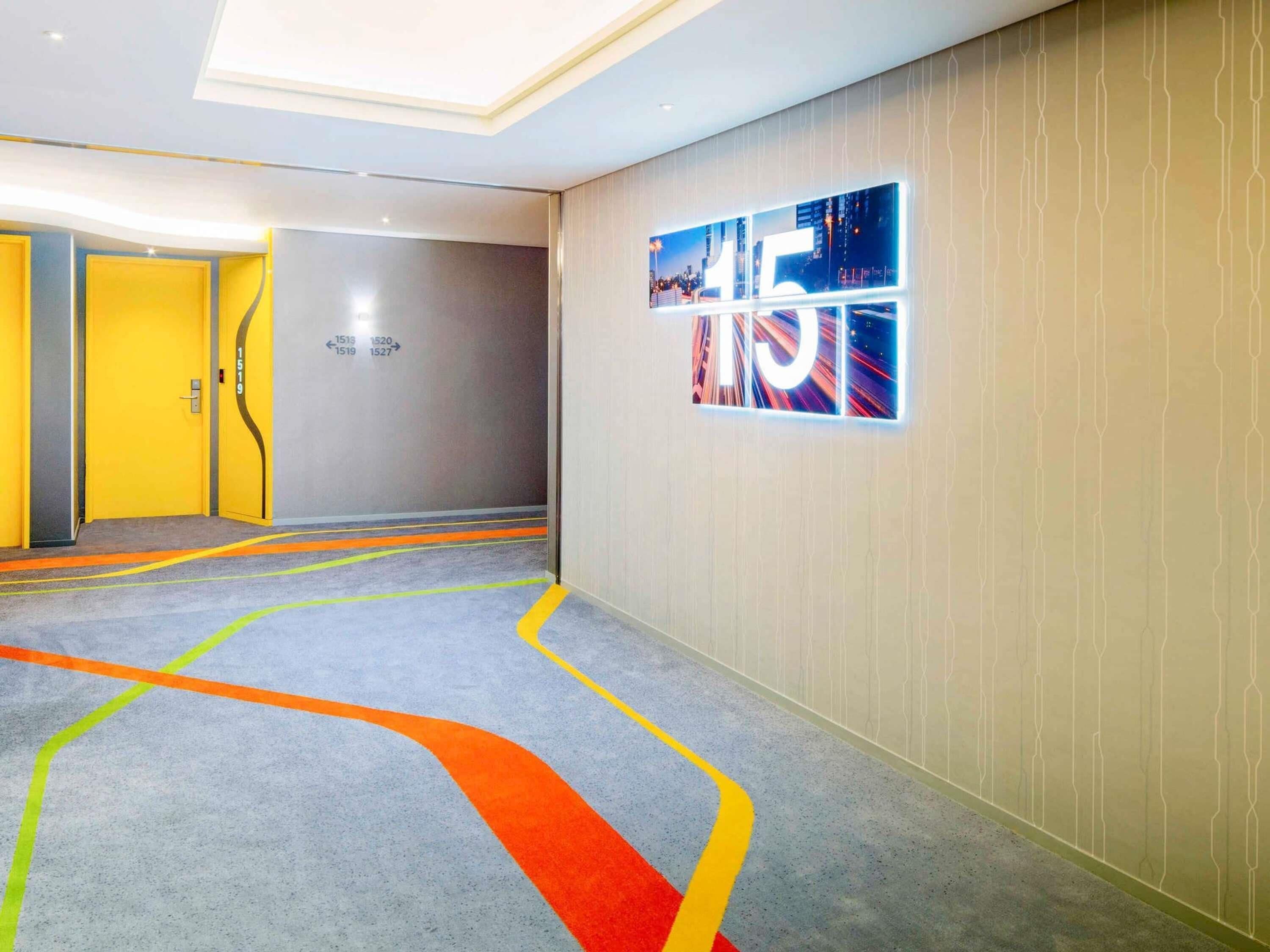 Corredor moderno de hotel com um painel digital na parede, marcações no chão indicando direção e uma porta com números de quarto.