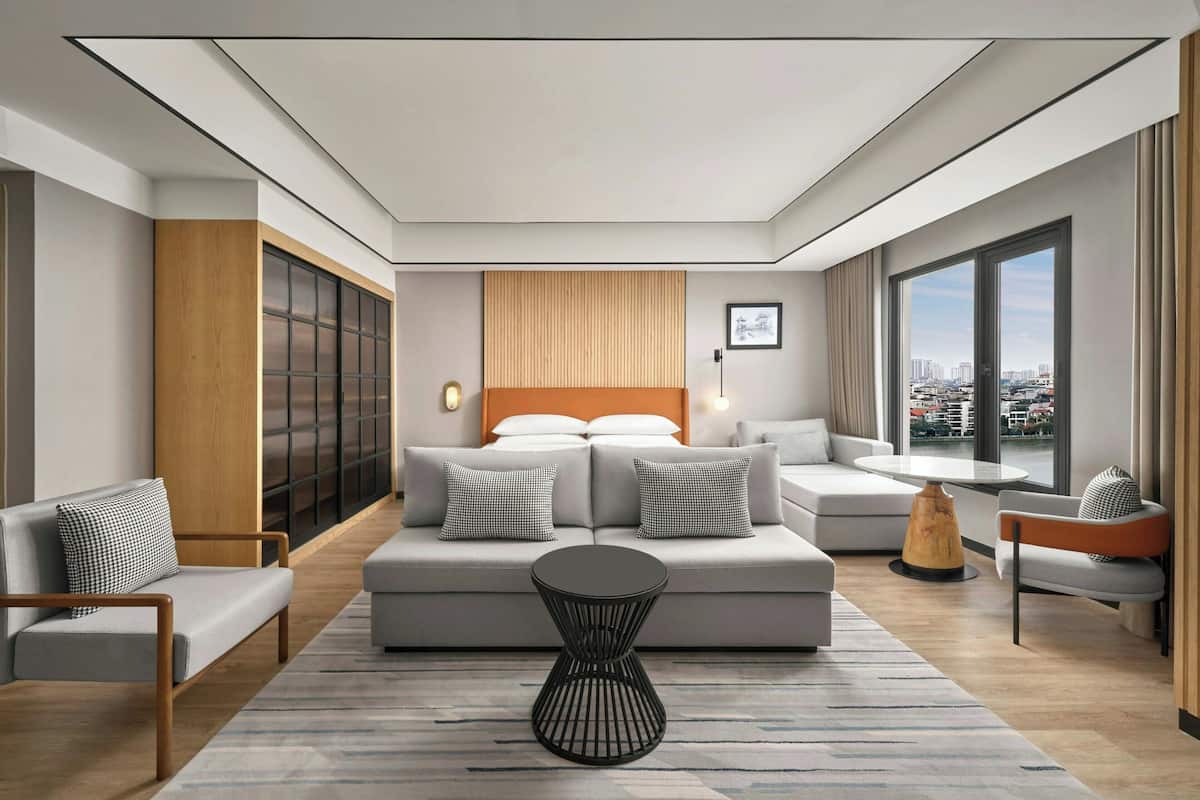 Quarto de hotel moderno com sofá, mesa de centro redonda, cama com cabeceira de madeira e uma ampla janela com vista para a cidade.