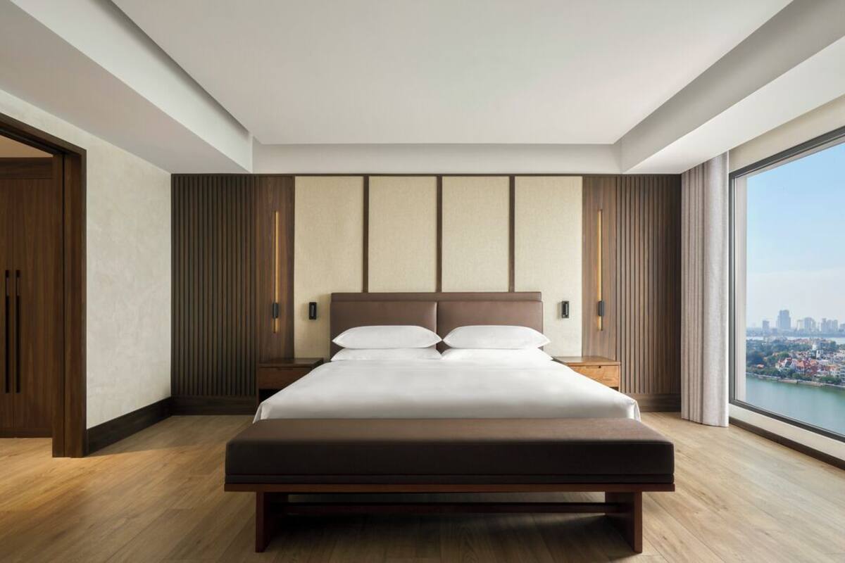 Quarto de hotel moderno com uma cama grande, vista para o horizonte urbano e piso de madeira.