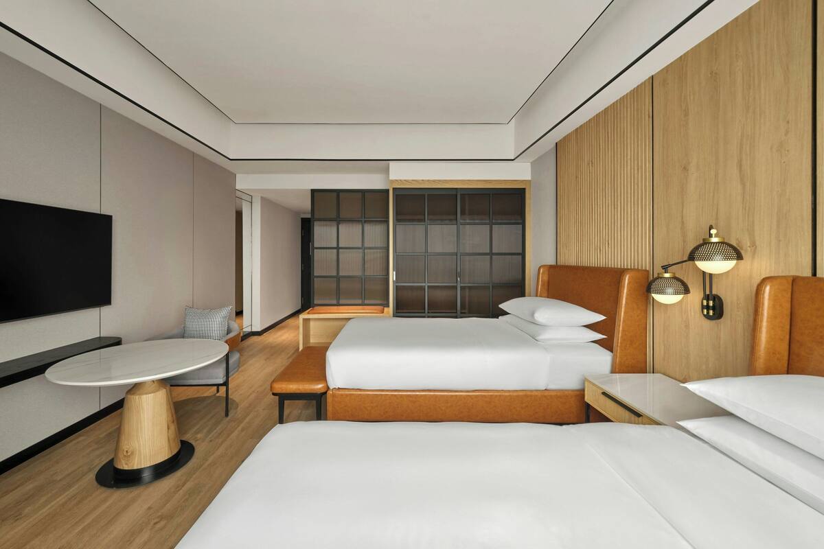 Quarto de hotel moderno com televisão de tela plana, duas camas, uma mesa pequena e um banco.