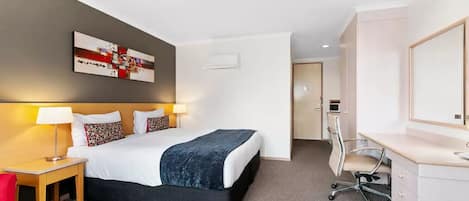 Quarto de hotel com cama, mesas de cabeceira, escrivaninha e uma cadeira. Há um quadro na parede e um aparelho de ar condicionado.