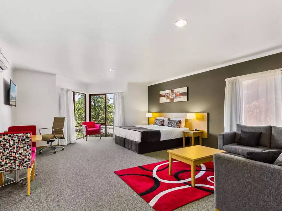 Quarto de hotel com cama, sofá, escrivaninha e cadeira. Há uma janela com cortinas, um tapete vermelho e um aparelho de ar condicionado na parede.
