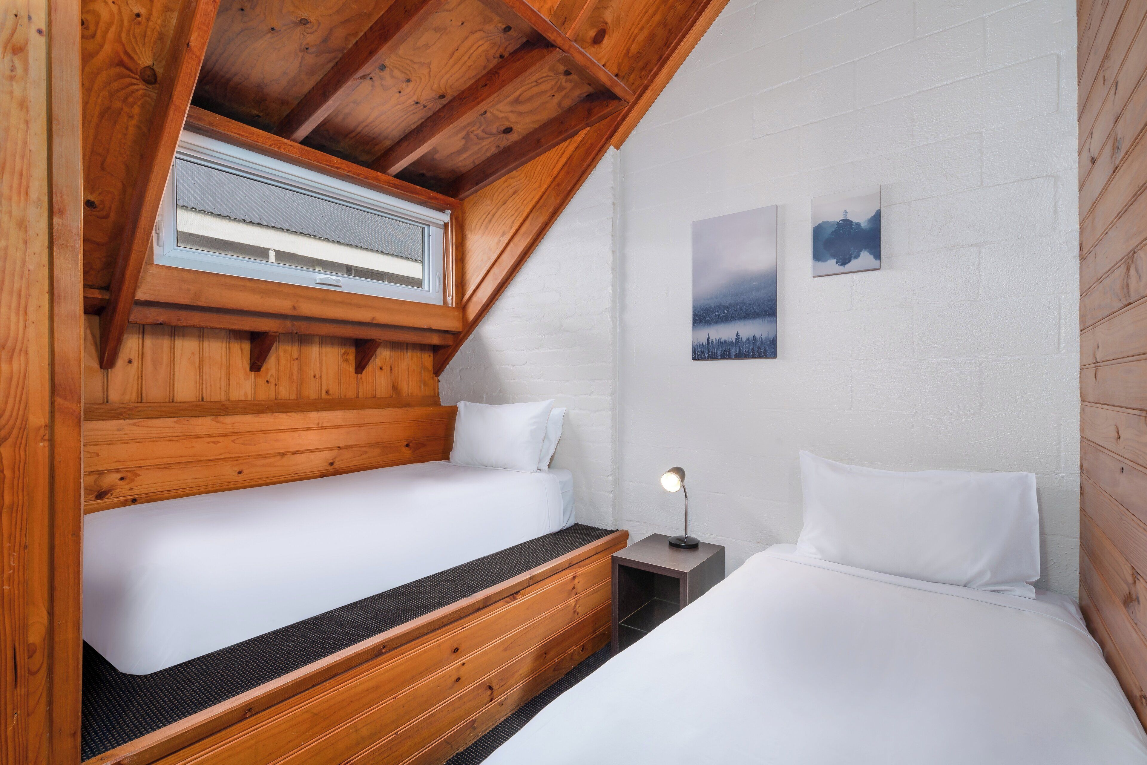Quarto com duas camas, vigas de madeira no teto, claraboia e paredes brancas.