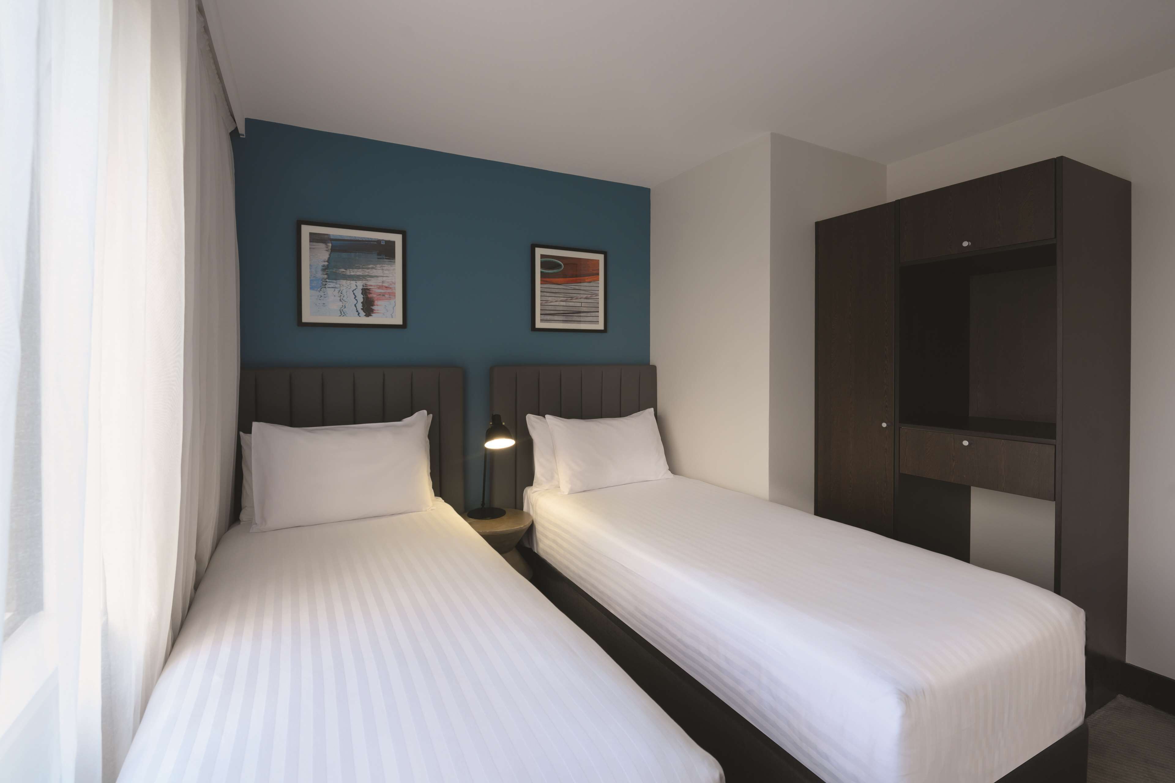 Quarto de hotel com duas camas de solteiro, mesa de cabeceira com abajur, guarda-roupa e duas fotografias emolduradas na parede.