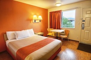 Quarto de hotel com cama, mesa de cabeceira, luminária, cadeira e janela com cortinas.