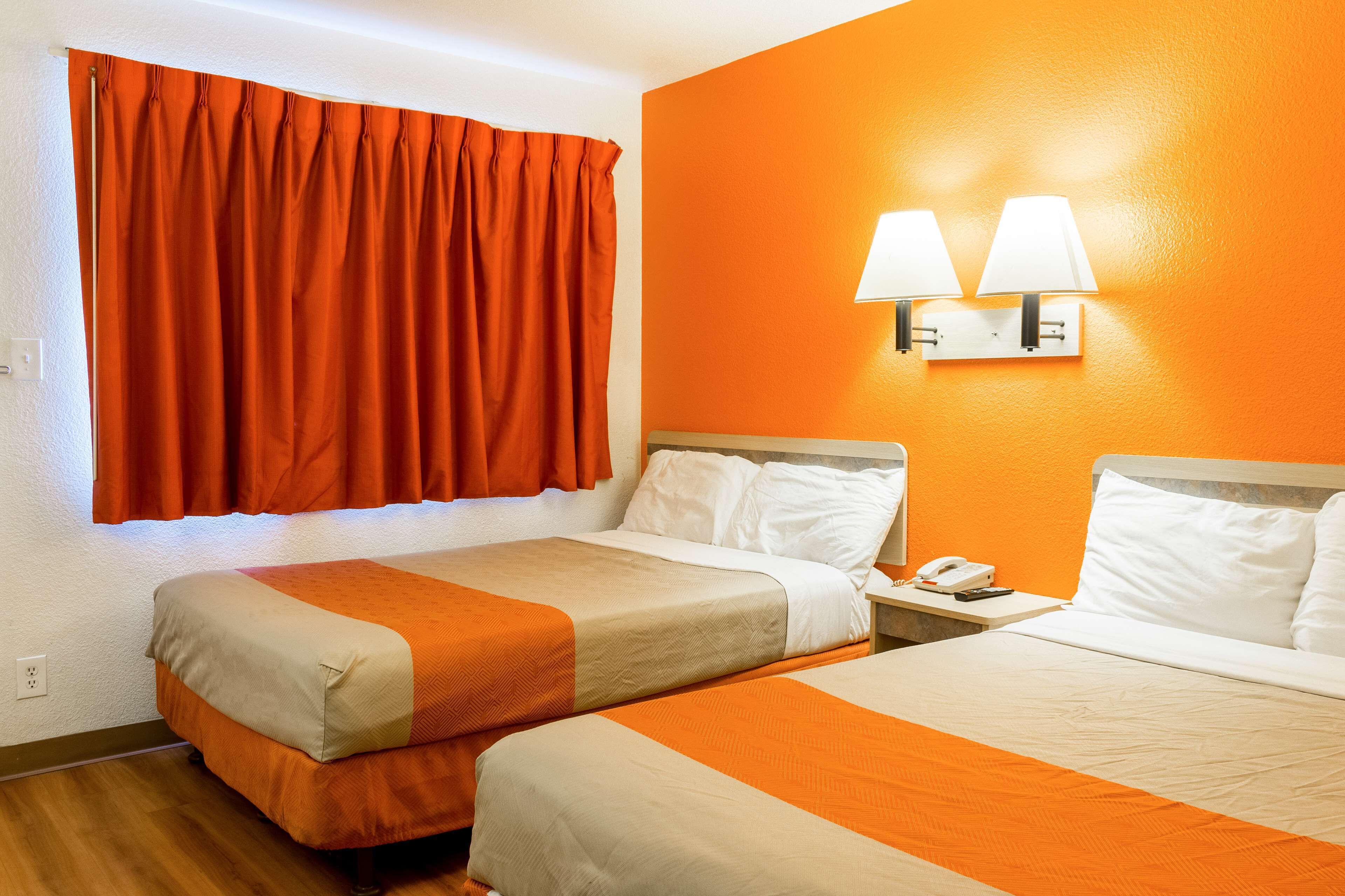 Quarto de hotel com duas camas, cortinas laranja e duas luminárias fixadas na parede.