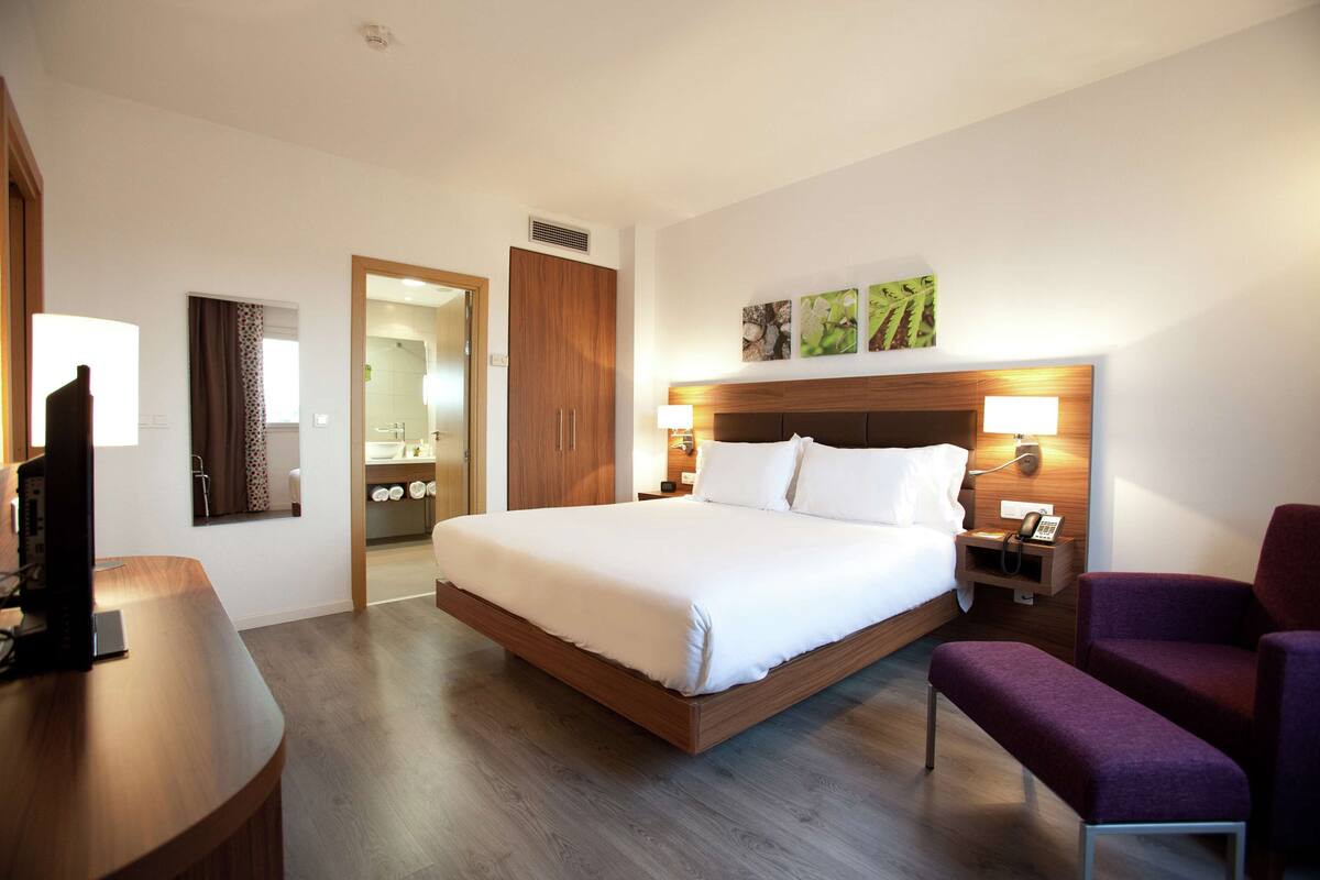 Quarto de hotel moderno com uma cama grande, uma escrivaninha, uma cadeira e um banheiro visível através de uma porta aberta.