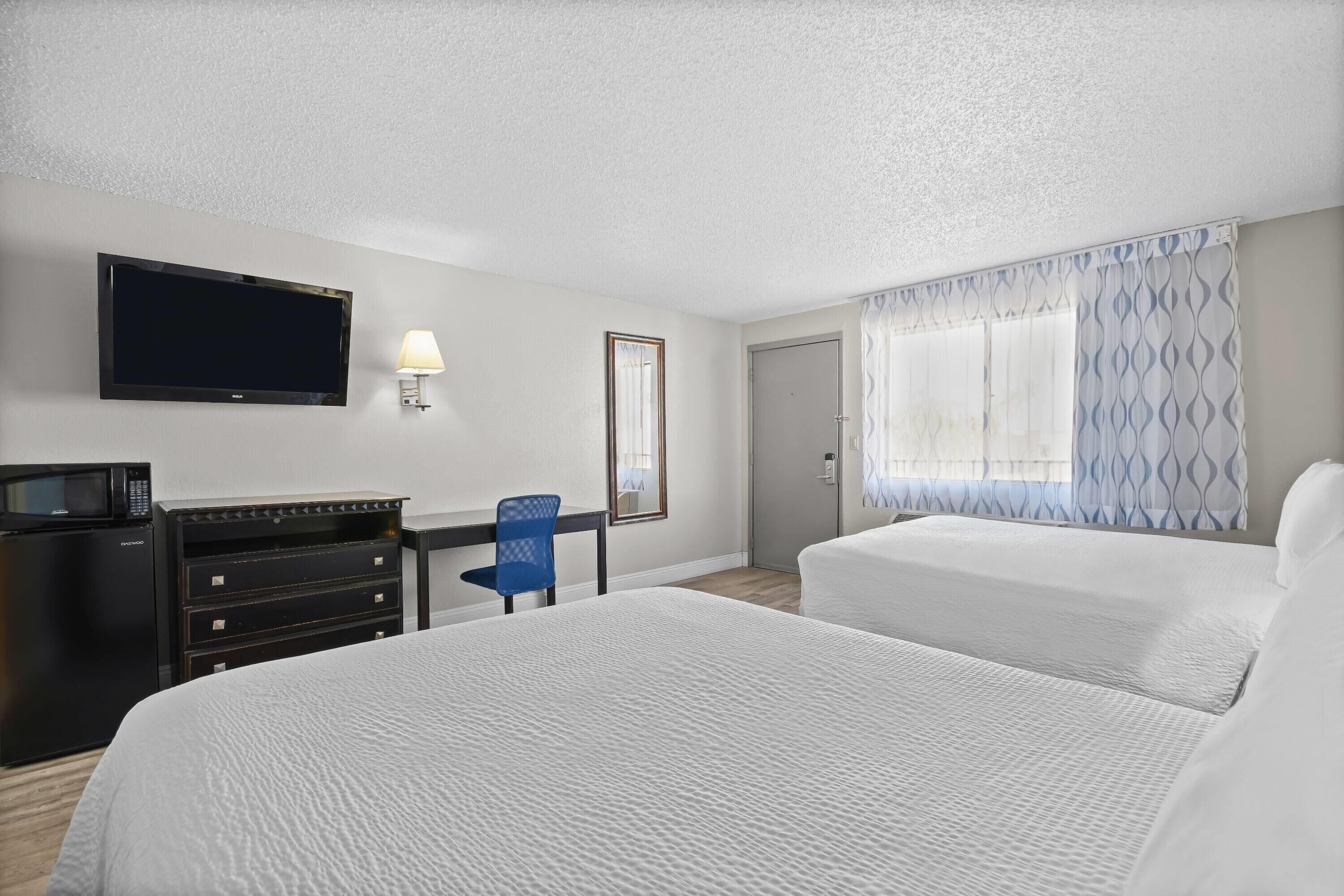 Quarto de hotel com duas camas, uma escrivaninha com cadeira, televisão, candeeiro e porta.