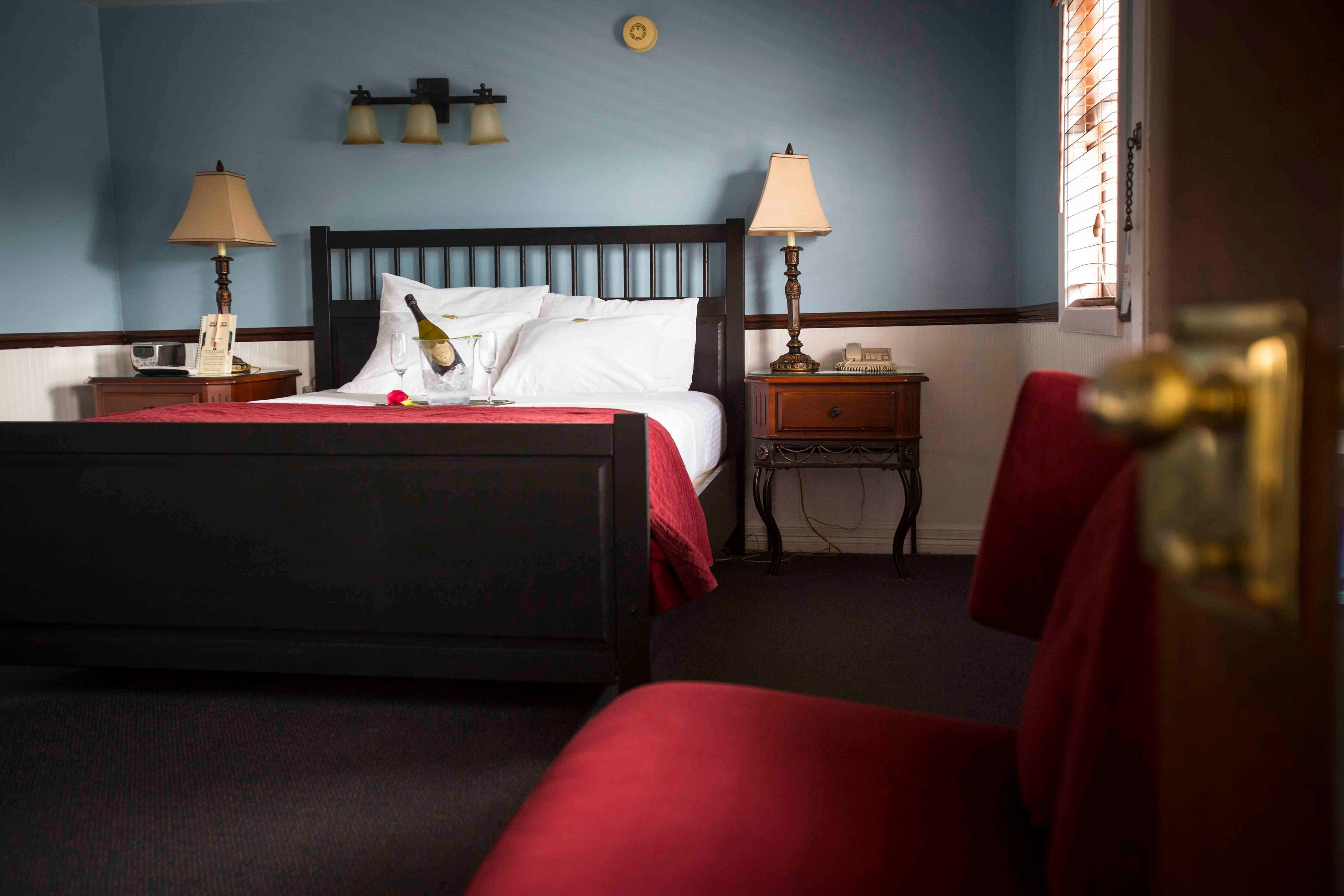 Quarto de hotel com cama, duas mesas de cabeceira, uma luminária e uma cadeira vermelha, além de uma janela com persianas.