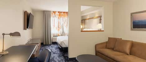 Quarto de hotel com cama, escrivaninha com cadeira, sofá, mesa pequena, televisão fixada na parede e um quadro na parede.