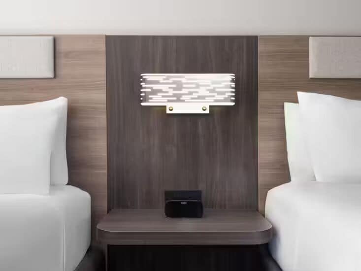 Quarto de hotel moderno com uma mesinha de cabeceira central, luminária fixada na parede e um pequeno dispositivo preto sobre a mesinha.