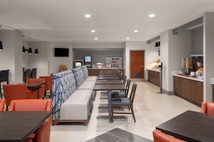 Uma área de refeições moderna com um longo banco, mesas individuais e cadeiras. Há um balcão com alimentos e uma televisão na parede.