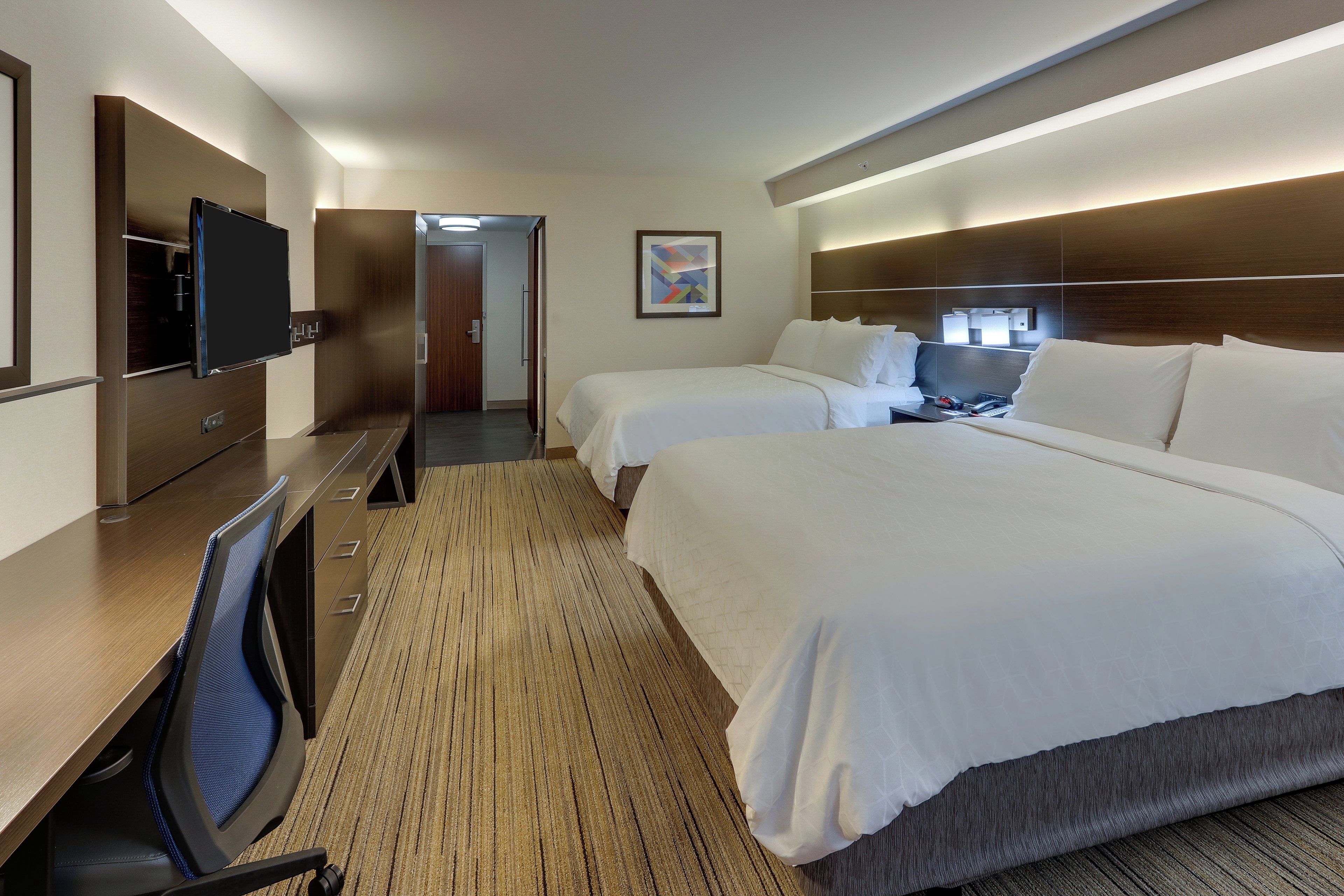 Quarto de hotel com duas camas, uma escrivaninha com cadeira, televisão e porta para um corredor.