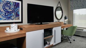 Quarto de hotel com escrivaninha de madeira, televisão de tela plana, micro-ondas, cadeira de escritório verde e uma janela grande.
