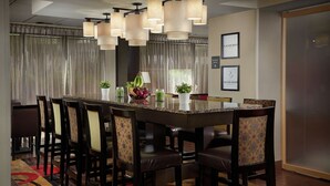 Uma área de refeições de hotel com uma longa mesa retangular, várias cadeiras e luminárias pendentes.
