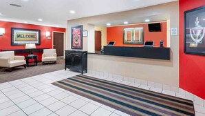 Uma área de recepção de hotel com parede vermelha, um letreiro 'WELCOME', balcão de recepção, televisão e uma moldura com imagem.