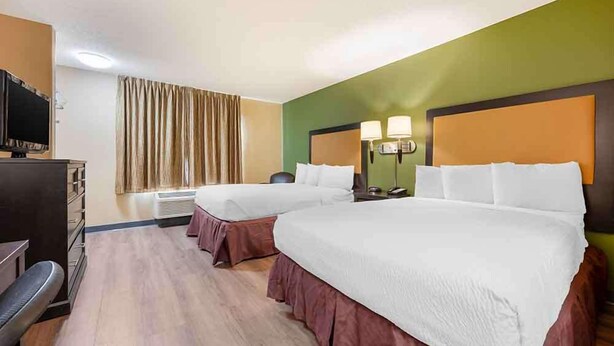 Quarto de hotel com duas camas, uma mesa de trabalho com cadeira, televisão e janela com cortinas.