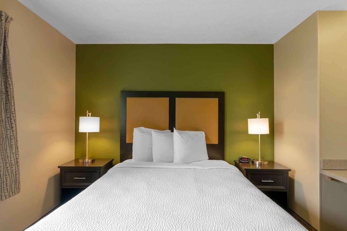 Quarto de hotel com uma cama grande, duas mesas de cabeceira com luminárias e uma parede verde de destaque.