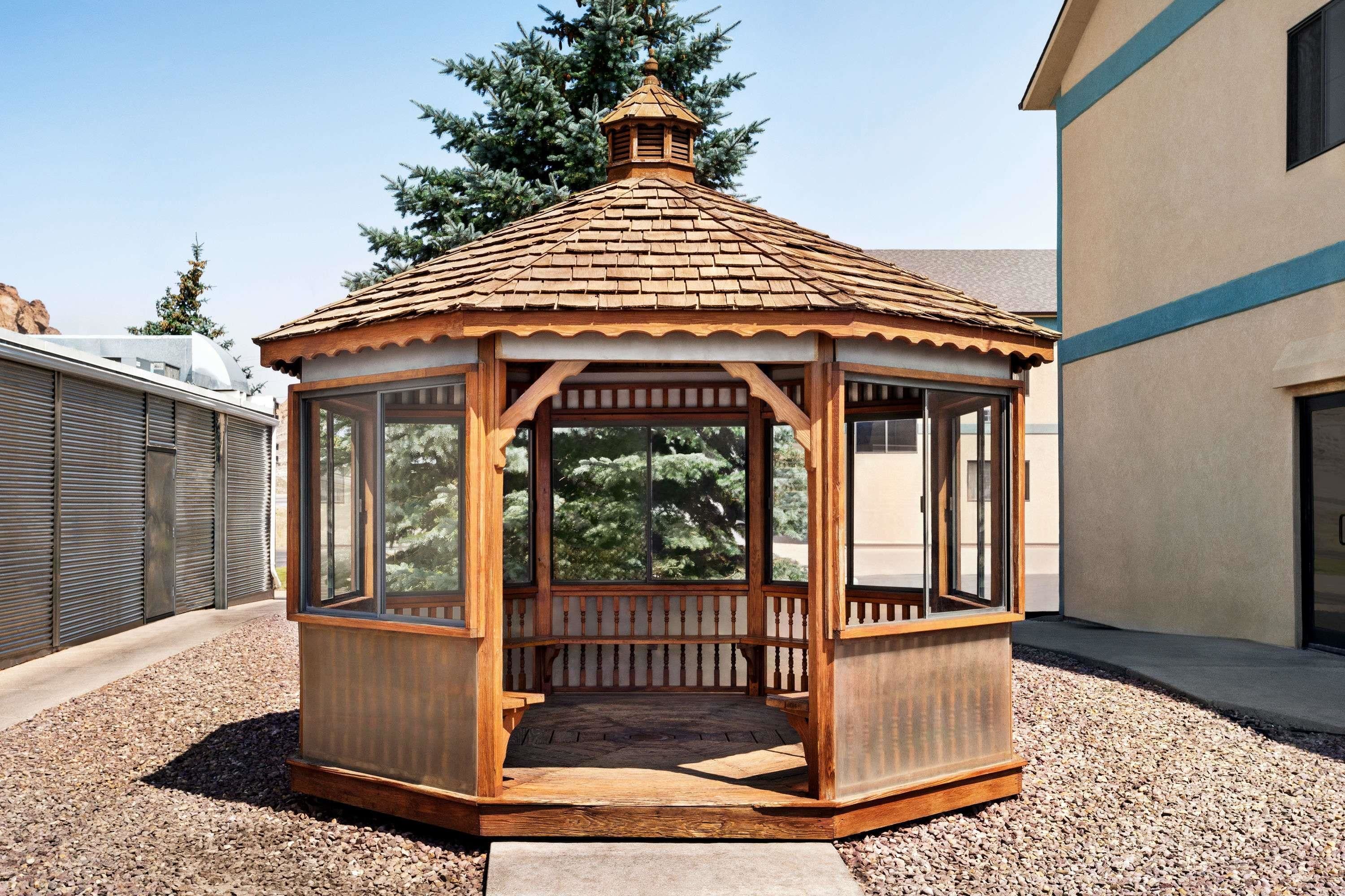 Um gazebo de madeira com telhado de telhas, rodeado por cascalho, e adjacente a um edifício com uma faixa azul.