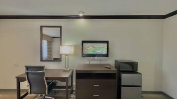 Quarto de hotel com escrivaninha, cadeira, televisão, luminária e pequeno frigorífico.
