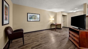 Quarto de hotel com chão de madeira, televisão de tela plana, uma cadeira, uma mesa com abajur e quadros nas paredes.