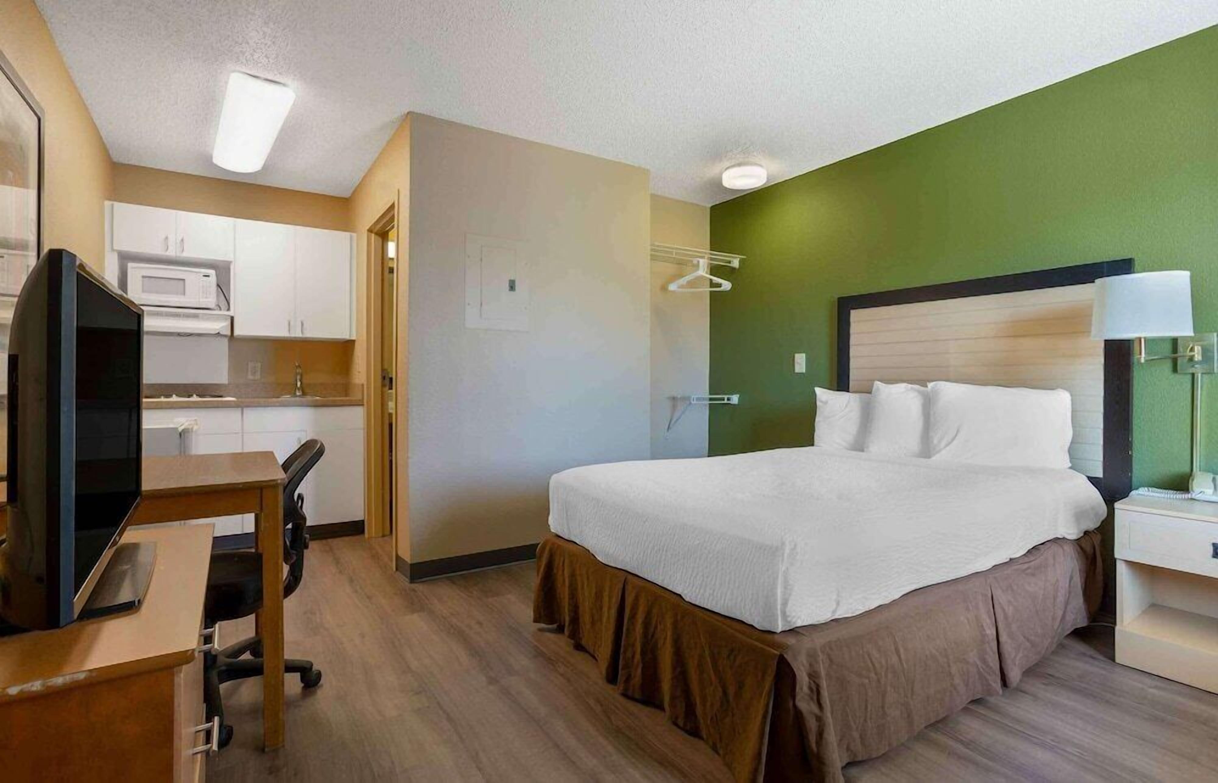 Quarto de hotel com cama, escrivaninha, TV, cozinha compacta e uma parede verde decorativa.