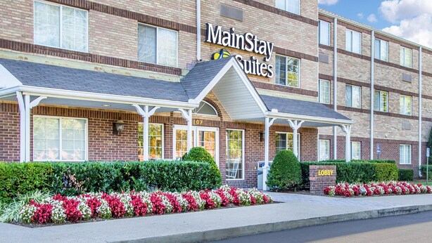 Um hotel MainStay Suites com fachada de tijolos, entrada coberta e um jardim bem cuidado com flores vermelhas e brancas.