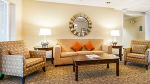 Um saguão de hotel com carpete estampado, um sofá com almofadas laranja, duas poltronas estampadas, uma mesa de centro e um espelho decorativo na parede.