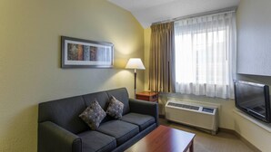 Quarto de hotel com sofá, televisão, luminária, janela com cortinas e um quadro na parede.