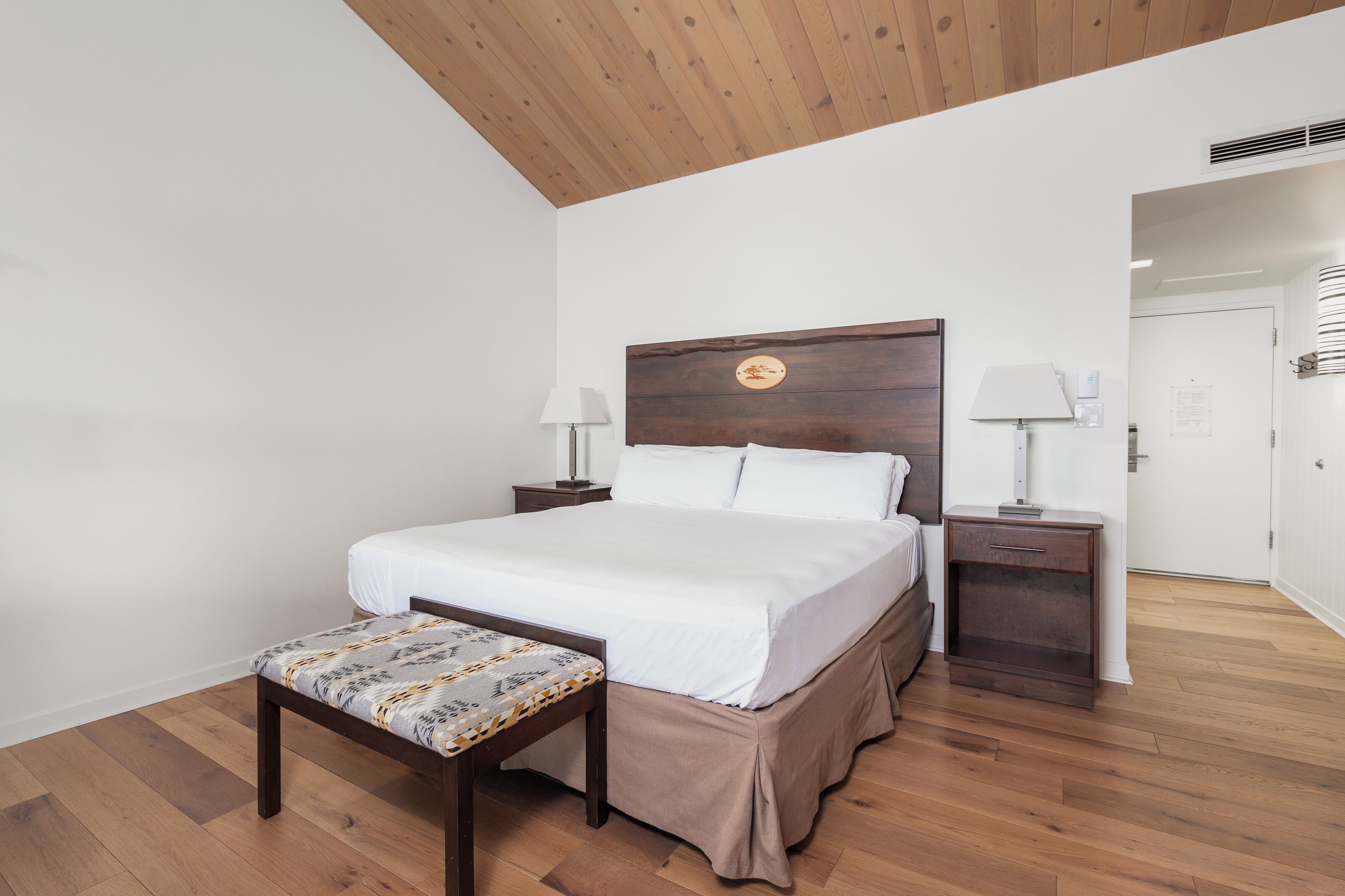 Quarto com teto de madeira, cama com roupa de cama branca, mesa de cabeceira com abajur e um banco com almofada estampada.