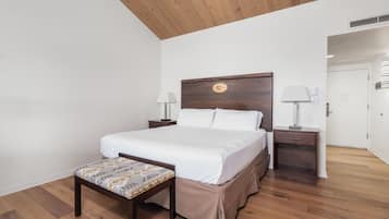 Quarto com teto de madeira, cama com roupa de cama branca, mesa de cabeceira com abajur e um banco com almofada estampada.